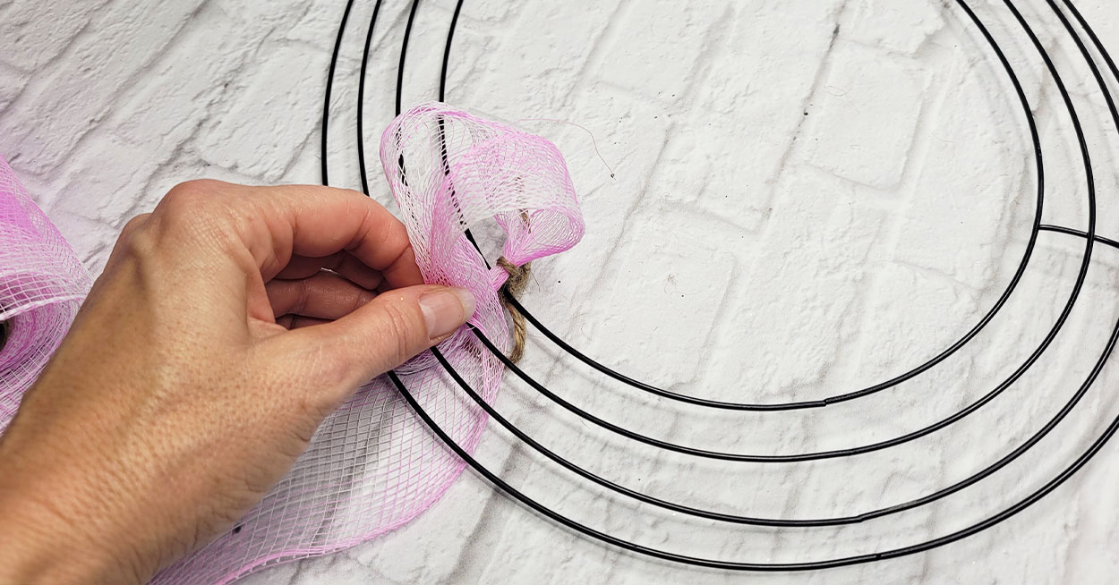 A hand holding pink decorative mesh tied to an egg-shaped wire wreath.
