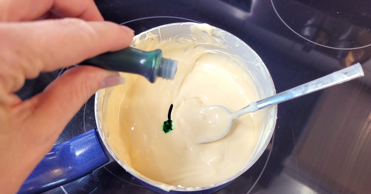 A hand dropping green food coloring into white chocolate in a pot on a ...