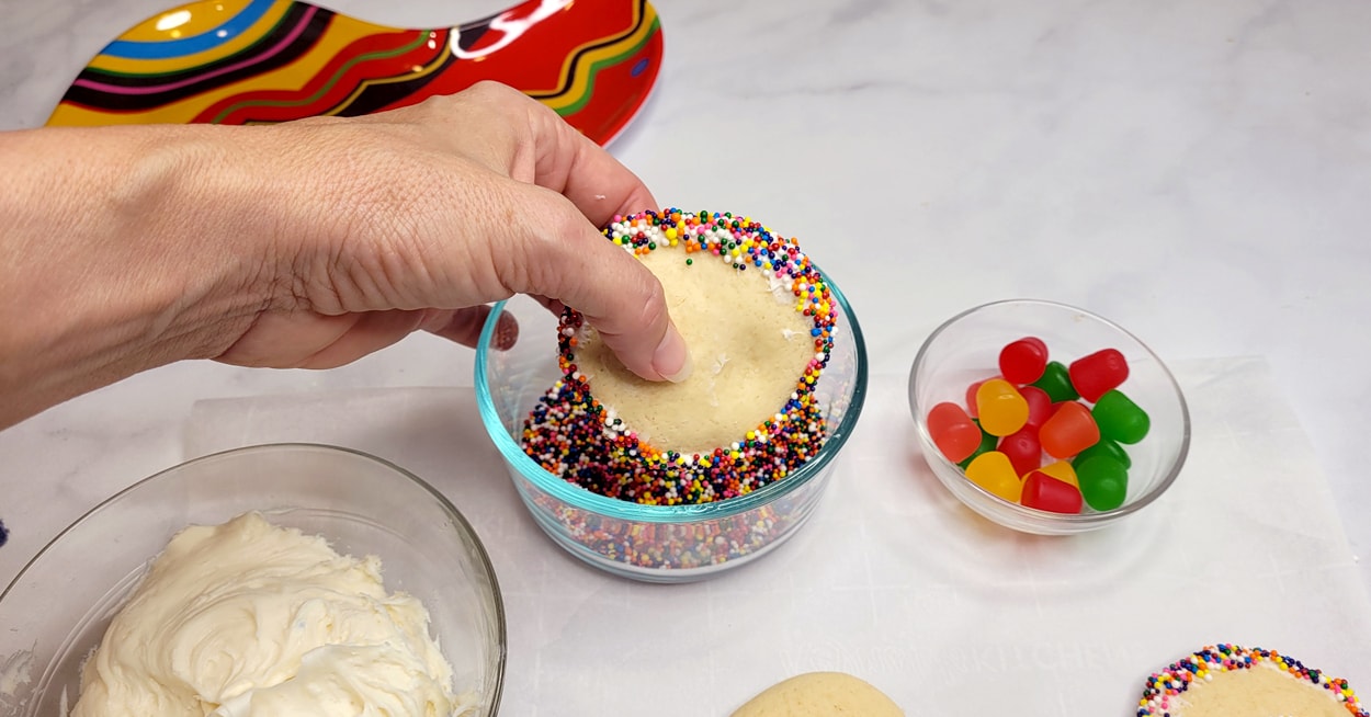 A hand dipping a cookie into sprinkles, gum drops, and a bowl of white ...