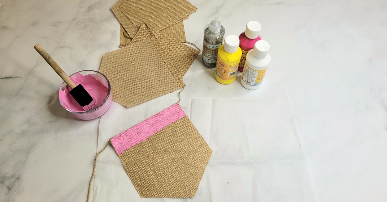 A burlap banner, a bowl of pink paint with a sponge brush in it, and ...