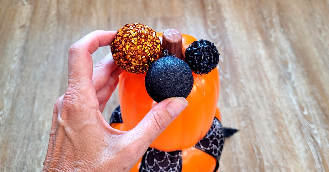 A hand holding glittery balls being glued on a pumpkin.