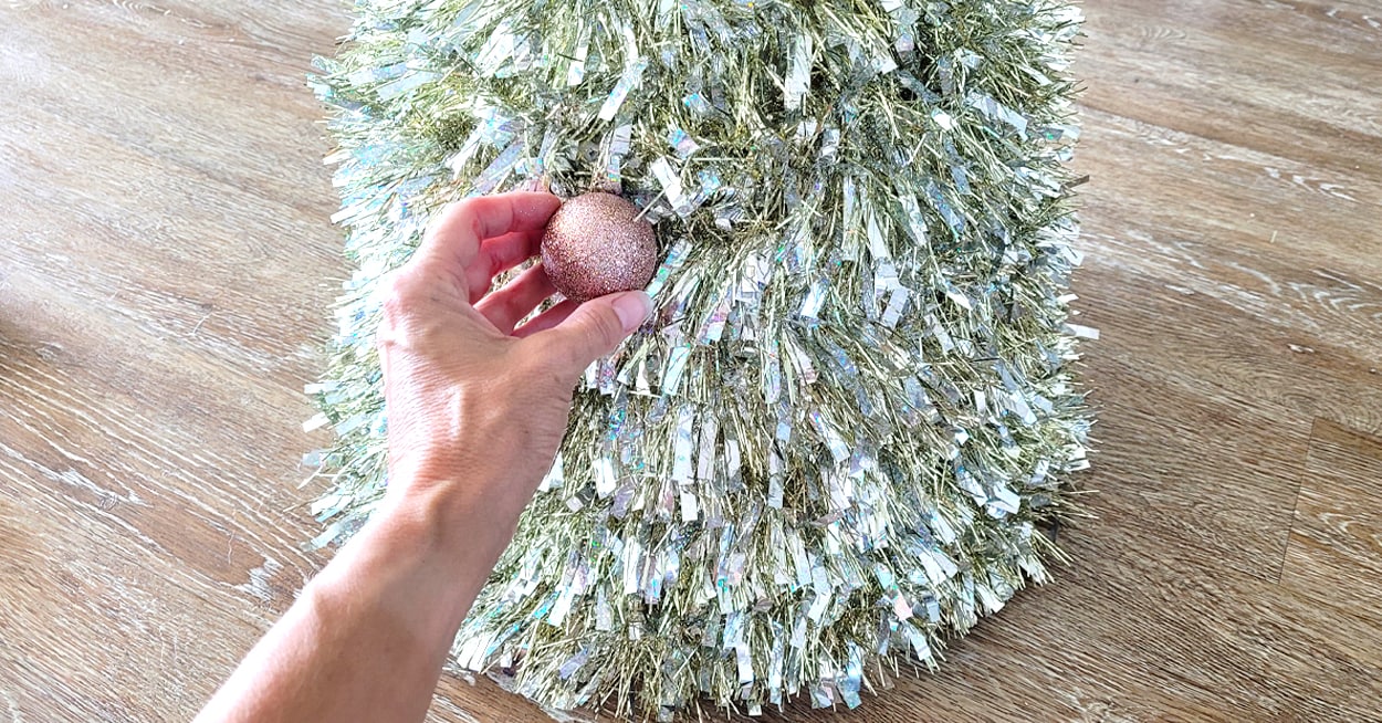 A hand holding a pink ball ornament onto a silver and gold tinsel tree.