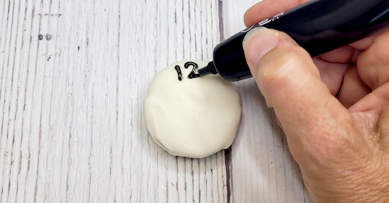 A hand holding a tube of black icing putting a number 12 on a white cookie.