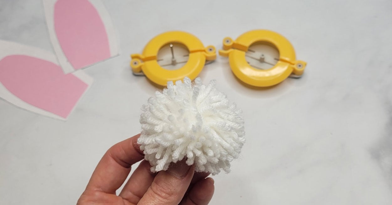A hand holding a white yarn pom pom with a pom pom maker and paper ...