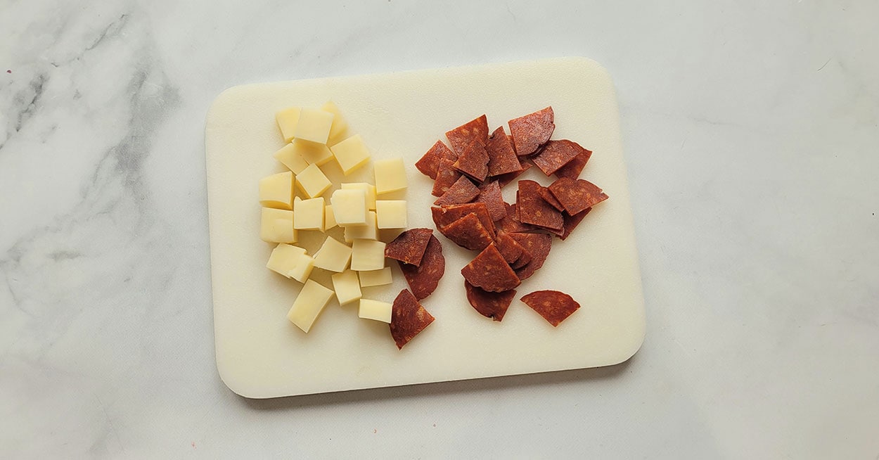 A cutting tray with cubes of white cheese and cut up pepperoni on it.