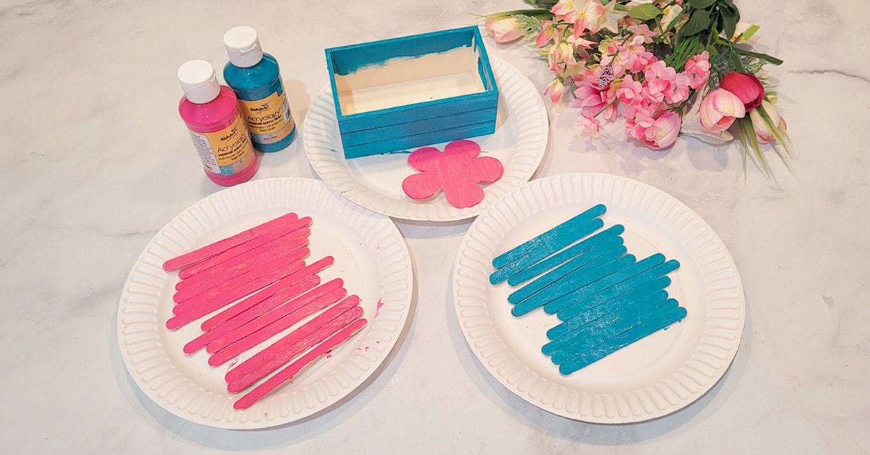3 white plates with pink and blue craft sticks, a small crate, pink ...
