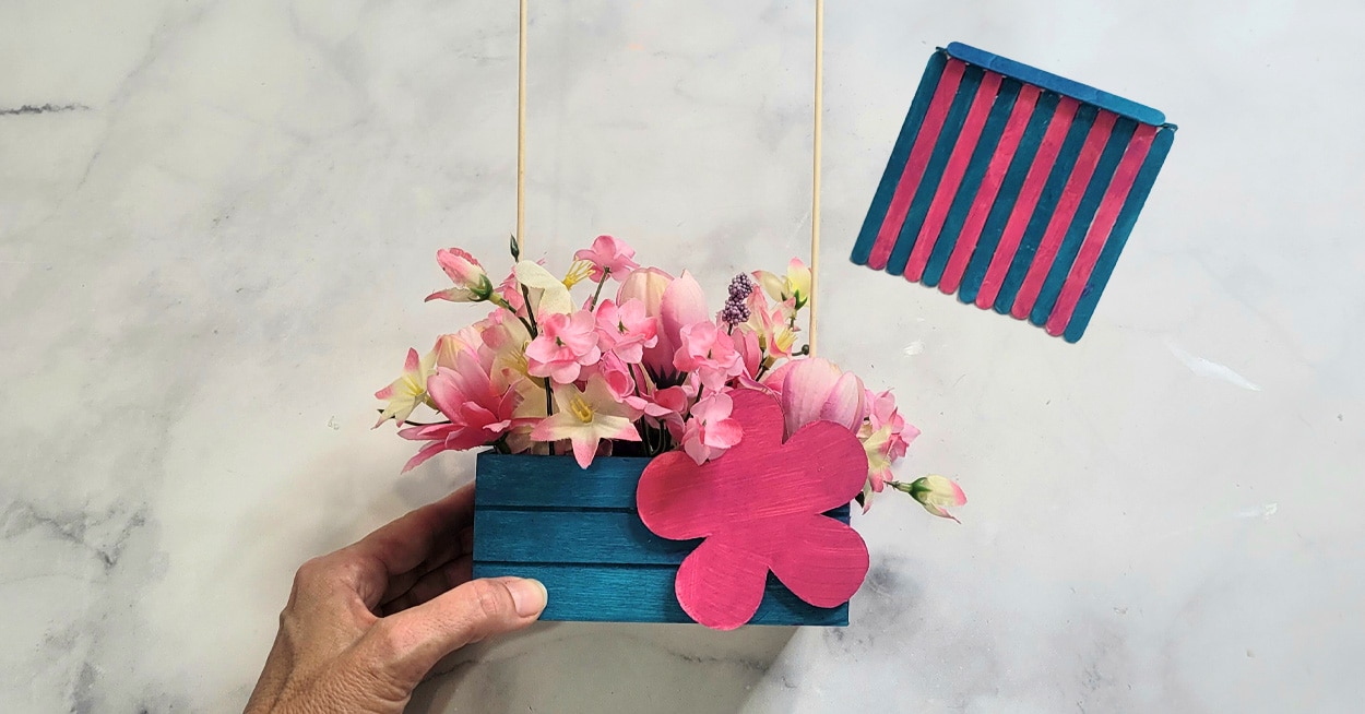 Hand holding a small blue wooden crate with skewers and flowers in it ...
