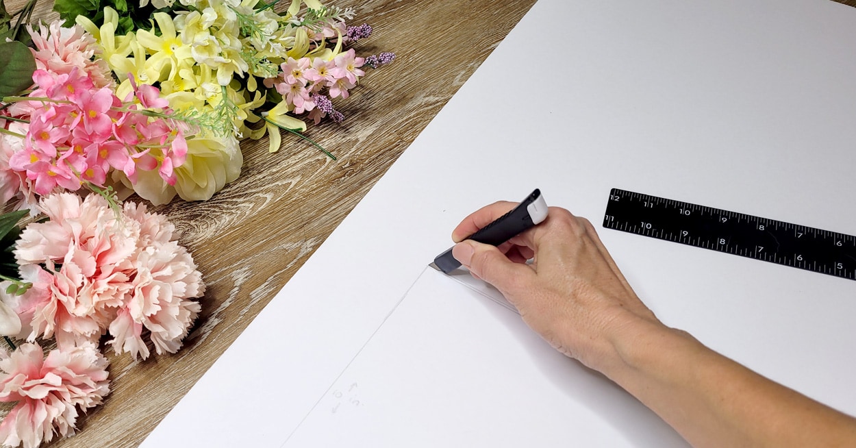 A hand holding a utility knife on a piece of white foam board, flowers ...